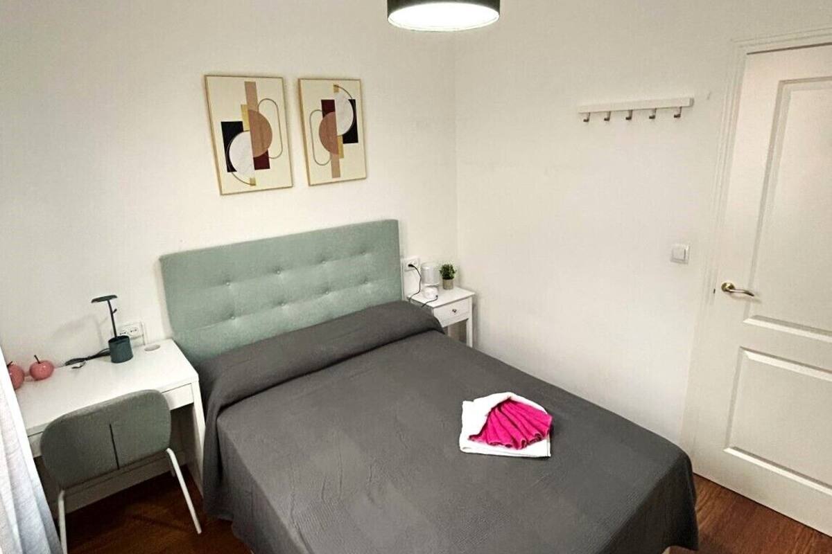 Quarto com cama, escrivaninha e cadeira. Há uma luminária no teto e uma porta.