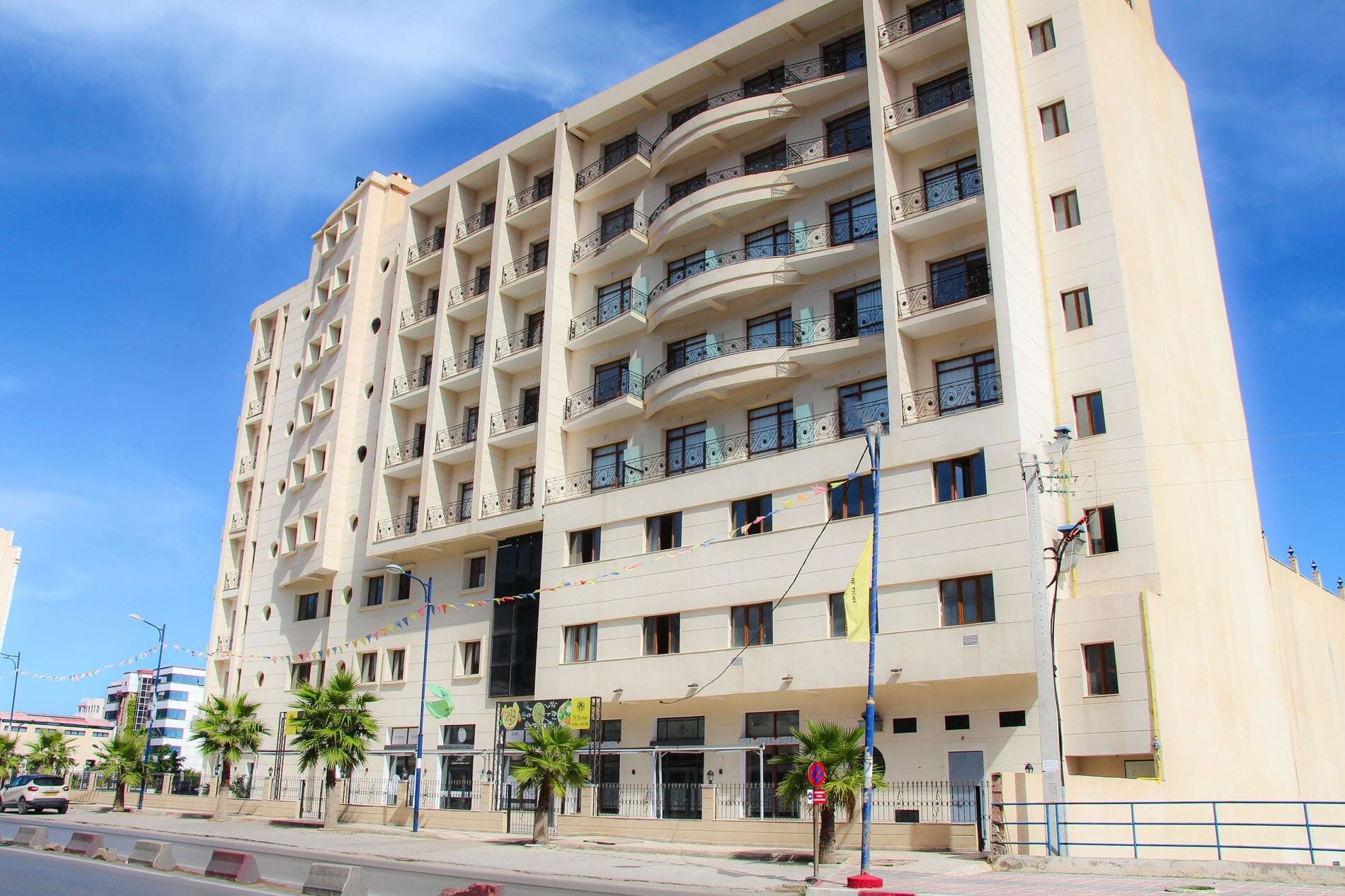 Um hotel de vários andares com varandas, entrada no nível do solo e palmeiras em primeiro plano.