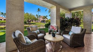 Uma área de estar coberta ao ar livre com móveis de vime e uma mesa central, com vista para um jardim paisagístico e um edifício ao fundo.
