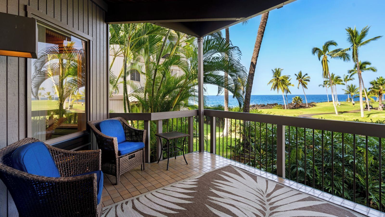 Uma varanda com cadeiras de vime e uma mesa pequena, com vista para uma paisagem tropical com palmeiras e o oceano.