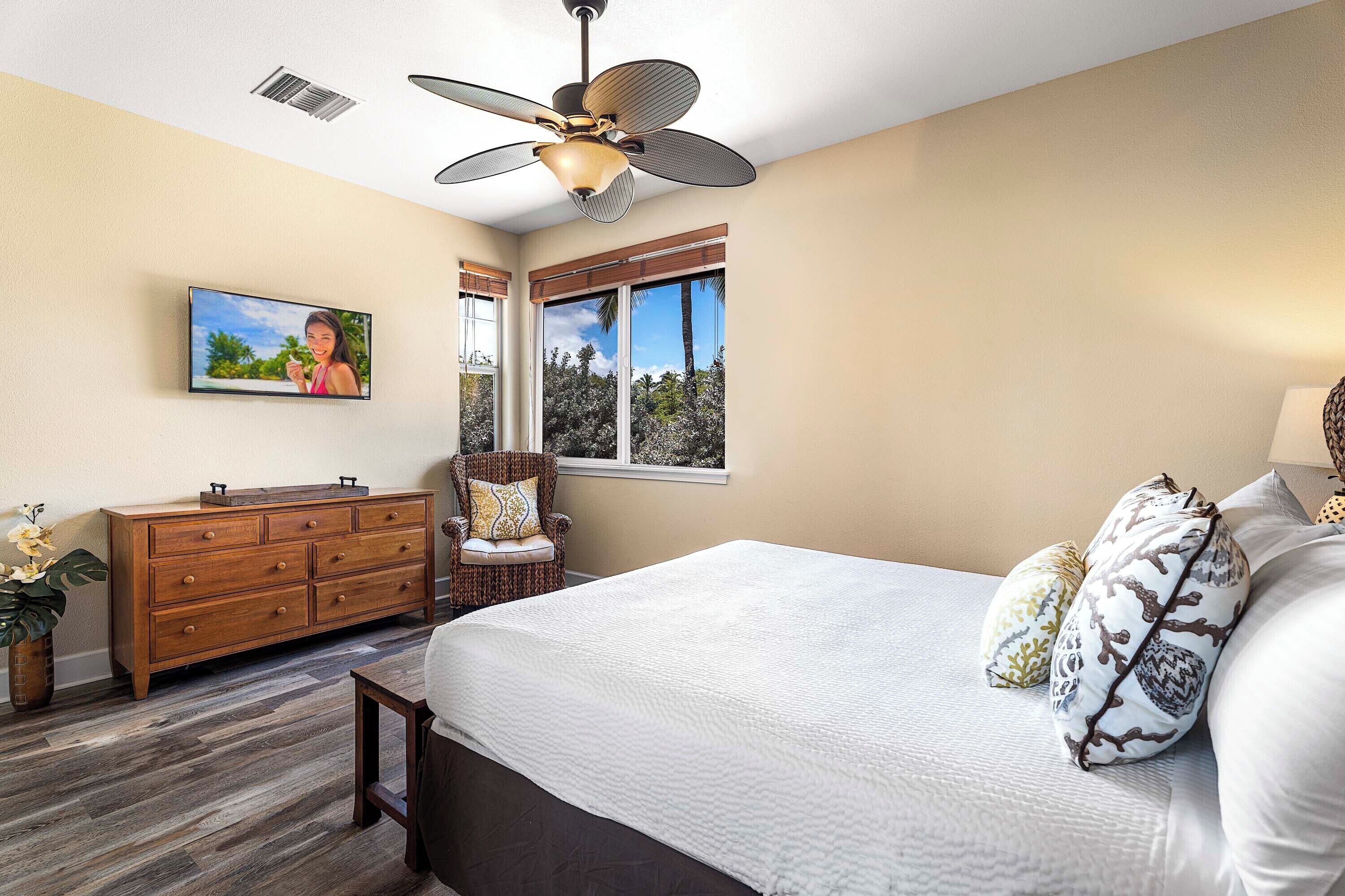 Quarto com uma cama grande, penteadeira, cadeira, ventilador de teto, TV e janela com vista para árvores.