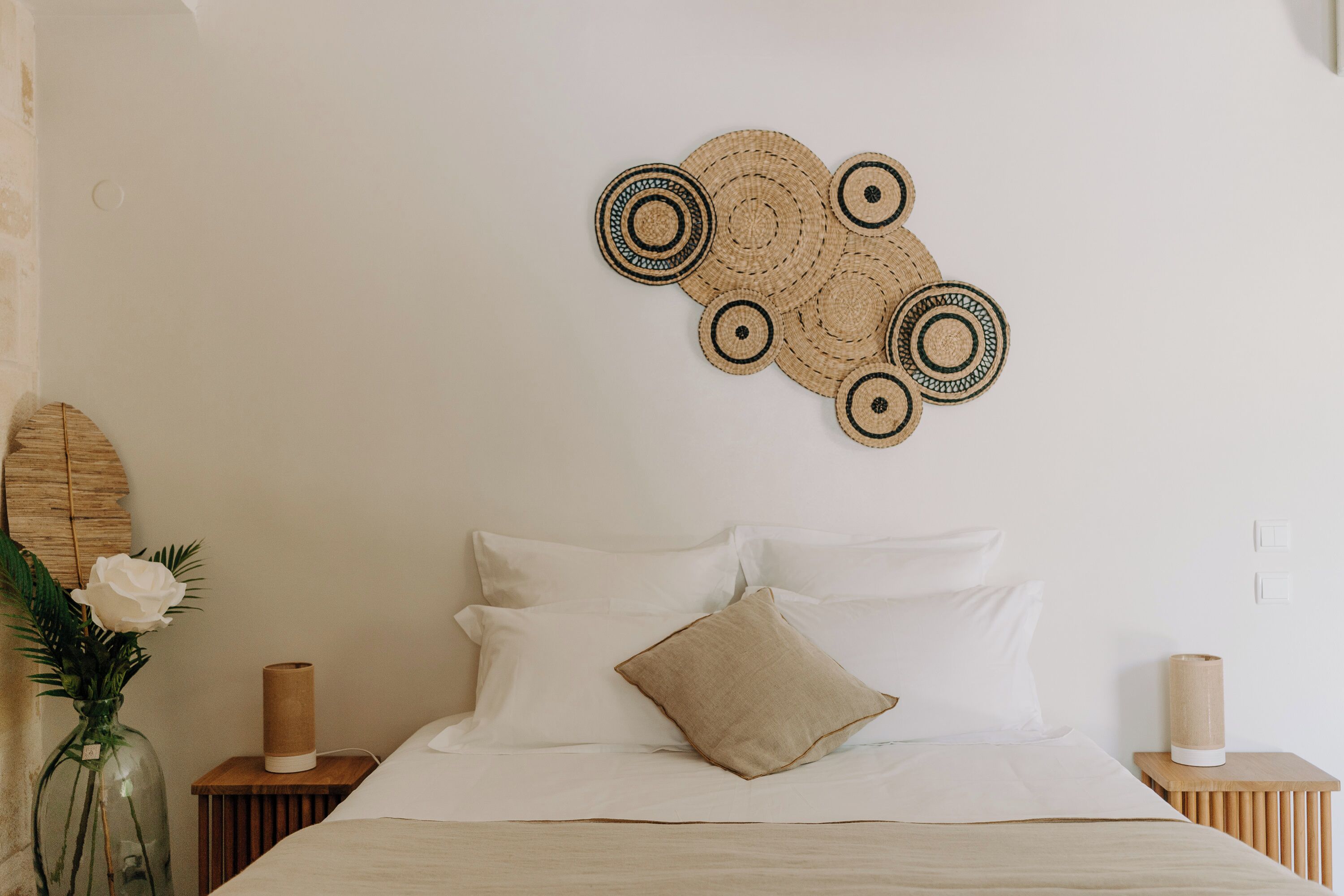 Quarto com cama, almofadas brancas, manta bege, um objeto decorativo na parede, um vaso com flores e uma mesinha de cabeceira de madeira com uma vela.