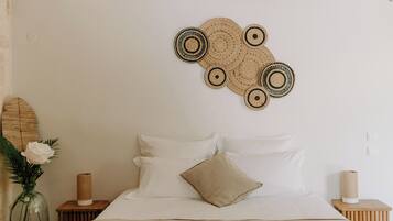 Quarto com cama, almofadas brancas, manta bege, um objeto decorativo na parede, um vaso com flores e uma mesinha de cabeceira de madeira com uma vela.