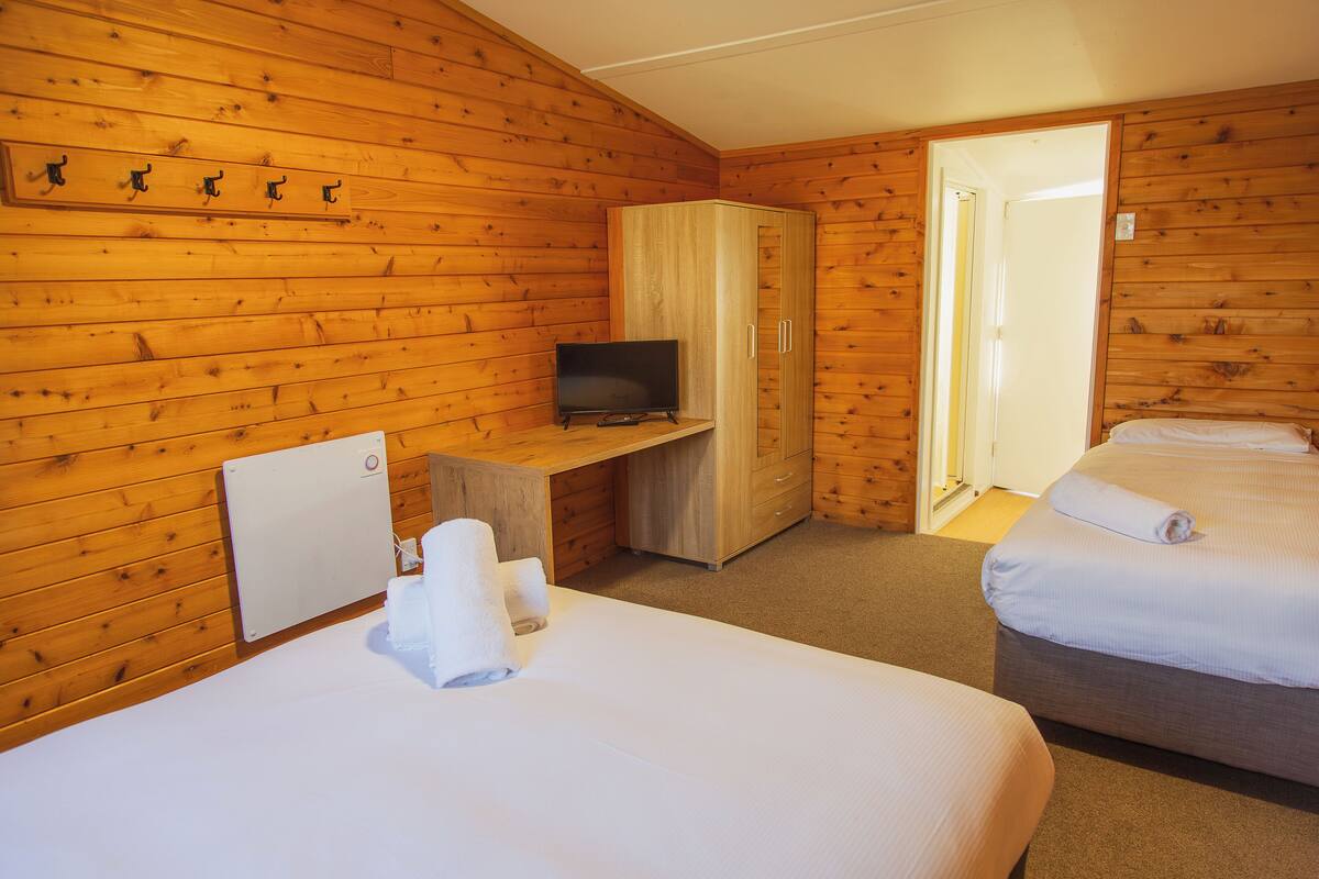 Quarto de cabana de madeira com duas camas, uma televisão sobre uma escrivaninha e um guarda-roupa.