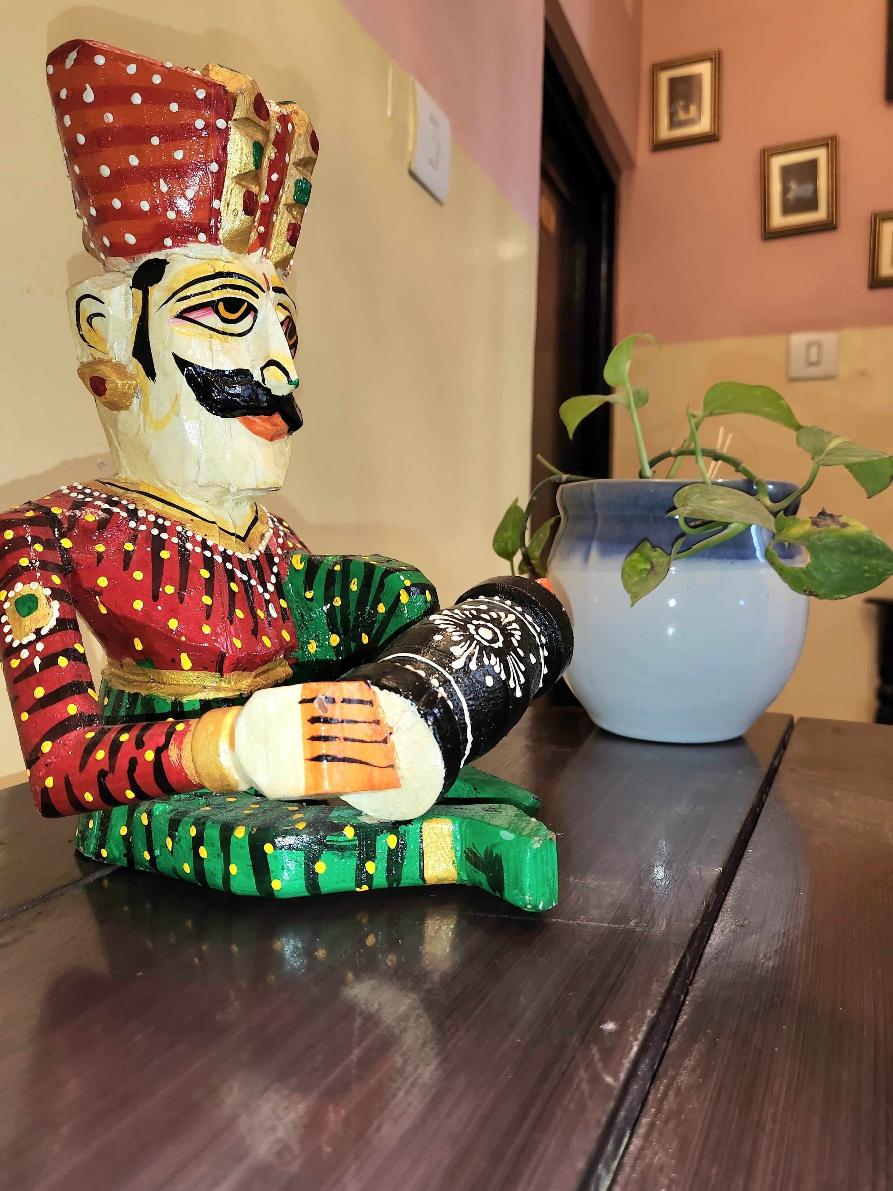 Uma escultura de madeira de uma figura usando chapéu vermelho e saia verde, colocada sobre uma mesa de madeira ao lado de um vaso azul com uma planta.