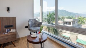 Quarto de hotel com uma grande janela, uma escrivaninha de madeira com telefone, uma poltrona estampada e uma mesinha com um vaso de flores.