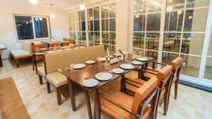 Uma área de refeições com uma mesa comprida posta para uma refeição, assentos acolchoados e grandes janelas com vista para a cidade.