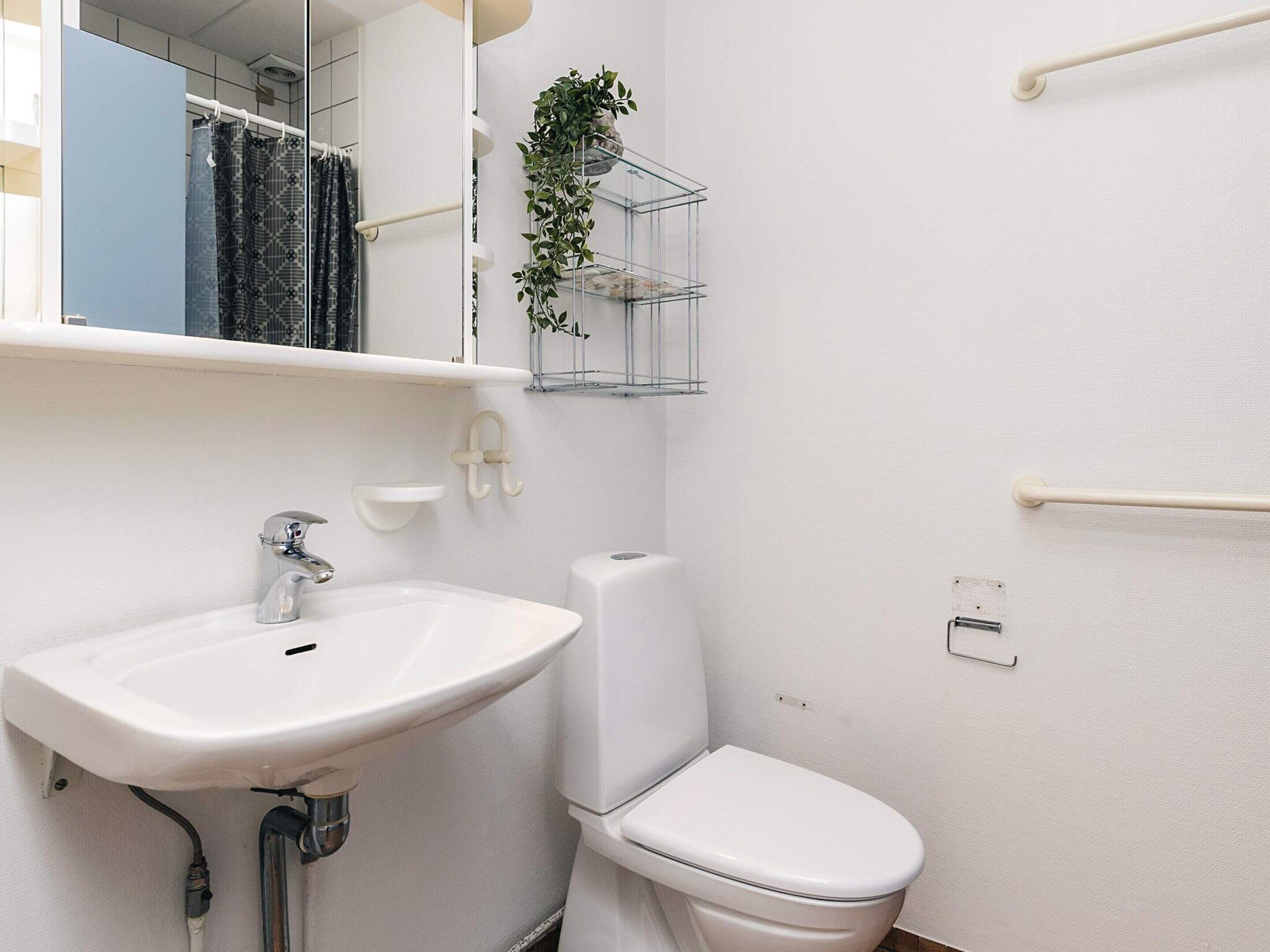 Uma casa de banho moderna com lavatório, sanita e cortina de chuveiro brancos. Há prateleiras com plantas e um porta-toalhas.