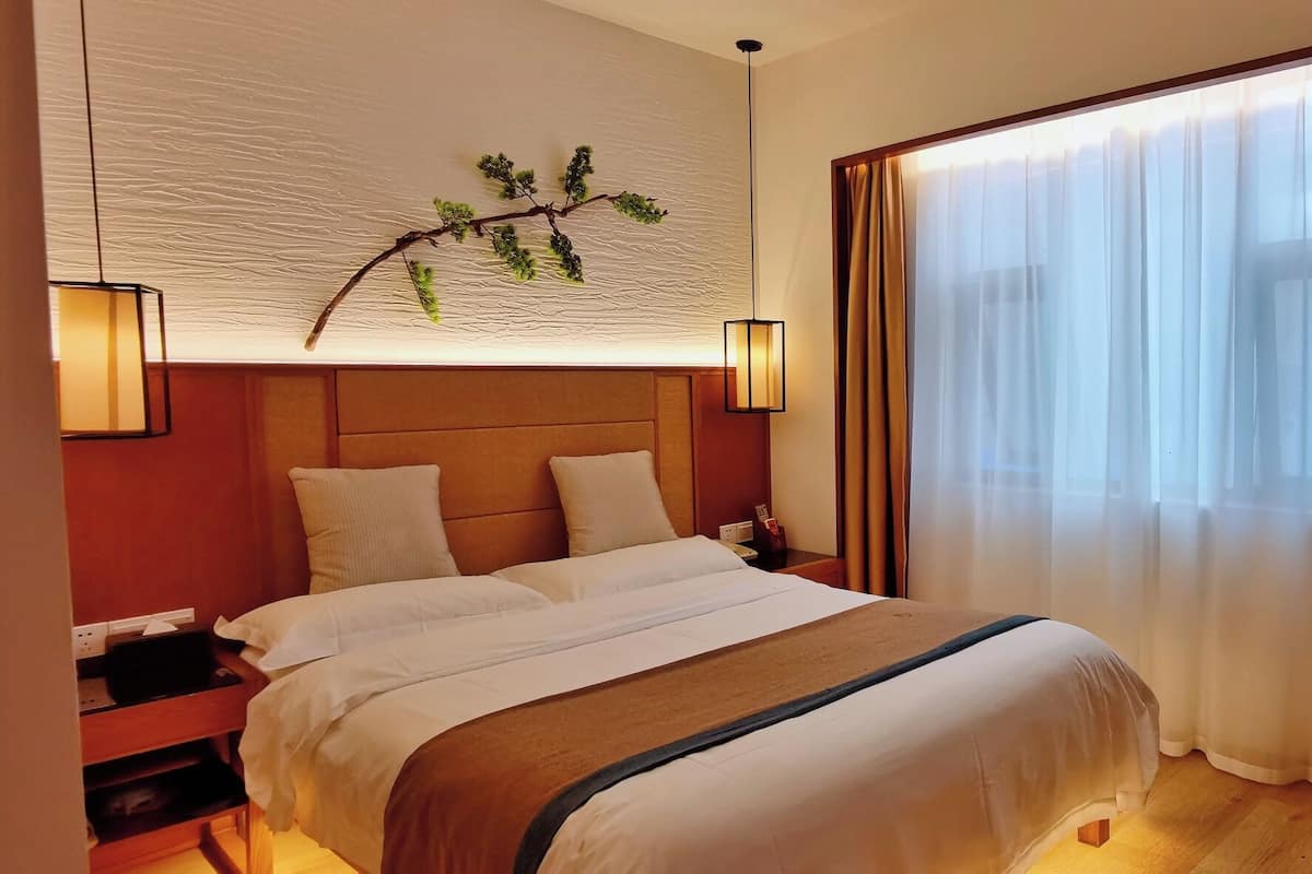 Quarto de hotel com cama, luminárias de cabeceira, planta suspensa na parede e cortinas de tule.