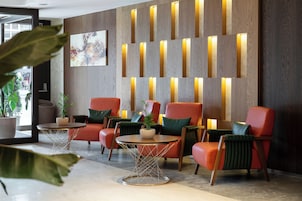Um hall com design moderno, destacando uma parede de madeira com painéis verticais de iluminação, uma mesa redonda com base de metal e várias poltronas com almofadas verdes e laranjas.
