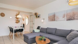 Uma sala de estar moderna com um sofá cinzento, uma mesa de centro redonda com uma planta, uma área de jantar com uma mesa e cadeiras brancas, e um espelho grande na parede.