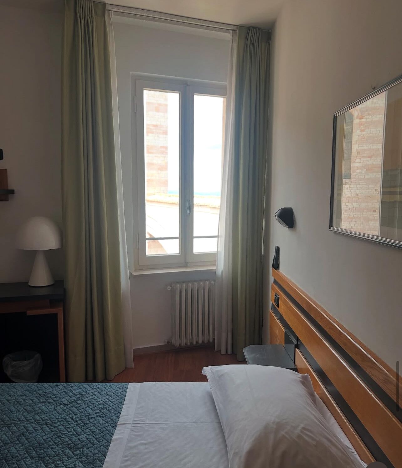 Quarto de hotel com cama, escrivaninha com luminária, janela com cortinas, radiador e um quadro na parede.