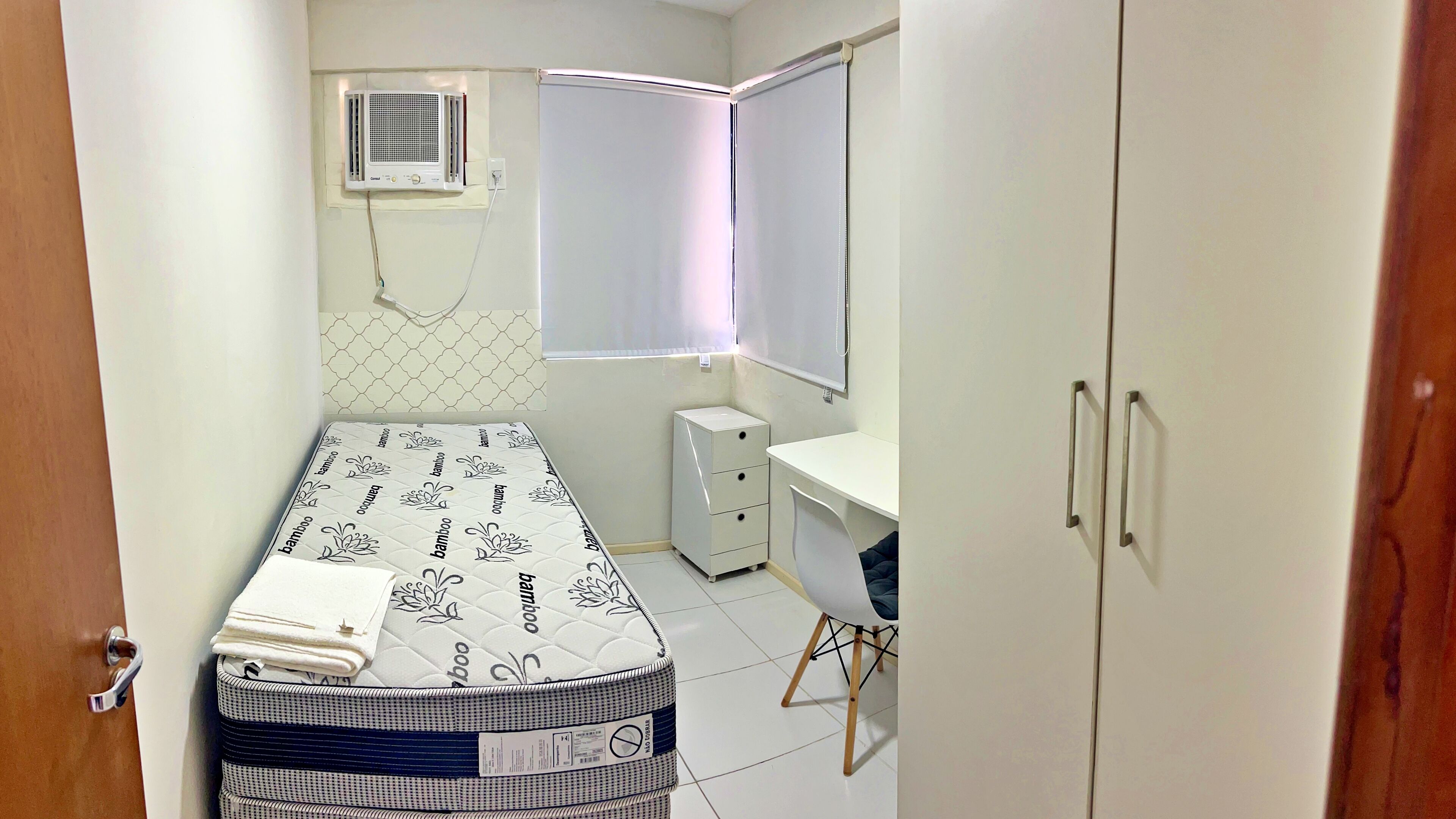 Um quarto pequeno e bem iluminado, com uma cama, escrivaninha, guarda-roupa e janela com persianas.