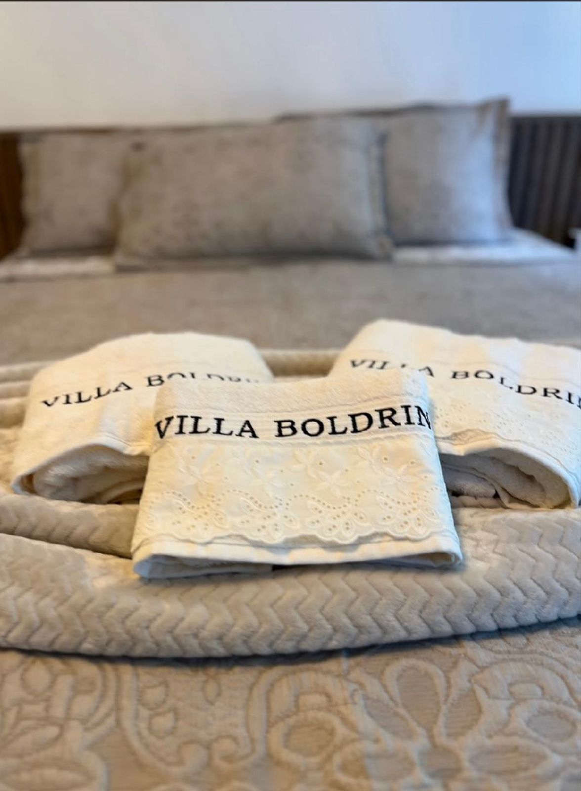 Cama com colcha bege estampada e duas toalhas dobradas com a inscrição "Villa Boldrin".