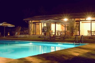 Uma área de piscina à noite, com um espaço coberto para sentar, espreguiçadeiras e uma piscina.