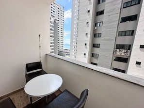 Varanda com mesa branca e duas cadeiras, com vista para edifícios altos e uma paisagem distante.