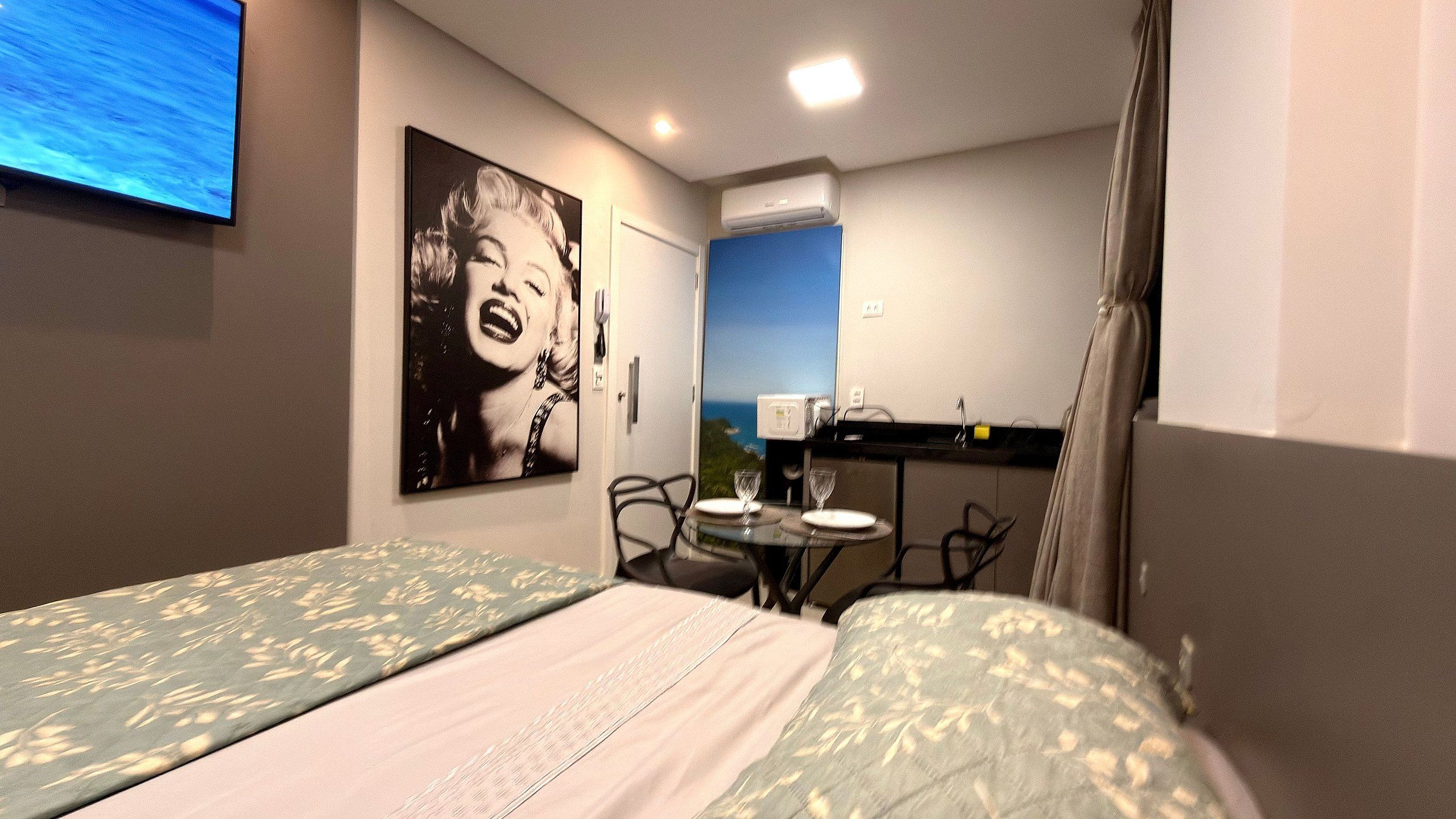 Quarto compacto com cama, uma mesinha com cadeiras, ar-condicionado na parede e televisão.