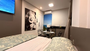 Quarto compacto com cama, uma mesinha com cadeiras, ar-condicionado na parede e televisão.