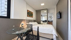 Quarto de hotel pequeno com uma cama, mesa de vidro, cadeira, televisão e um quadro na parede.