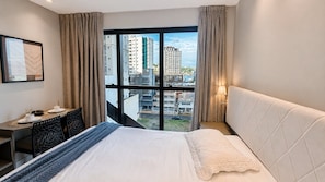 Quarto de hotel com uma cama grande, uma mesa de trabalho e vista para a paisagem urbana através de uma janela ampla.