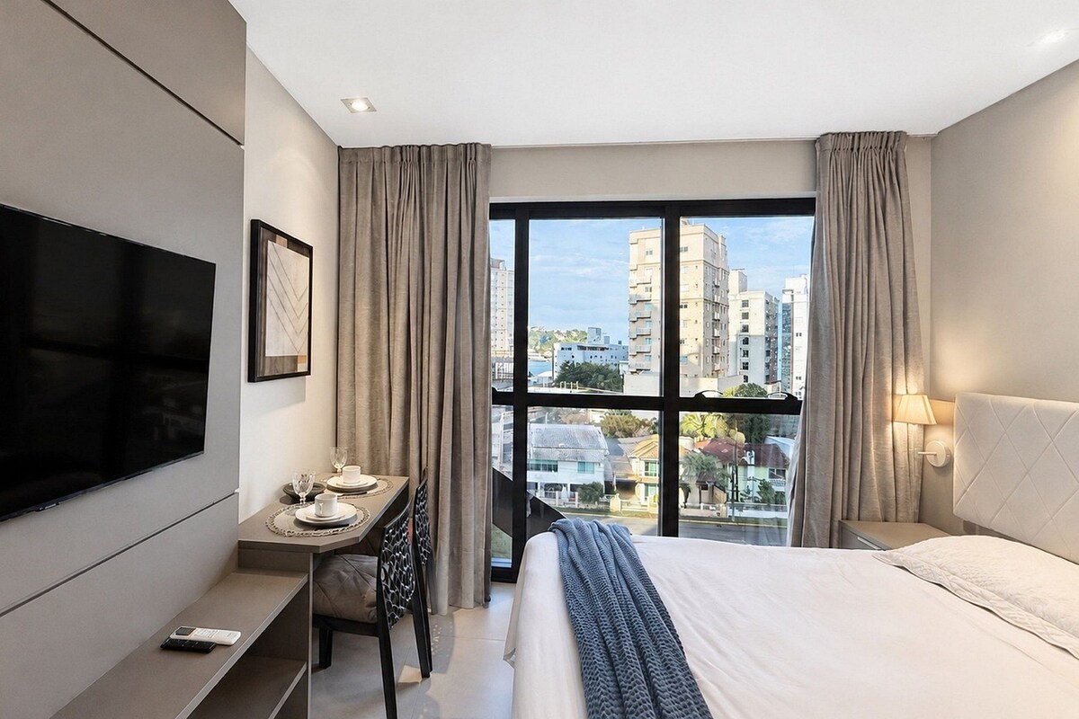 Quarto de hotel com cama, escrivaninha, televisão e janela com vista para a cidade.