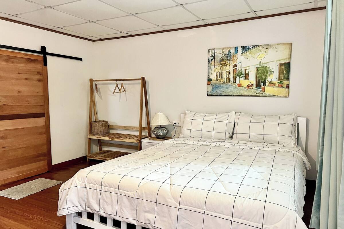 Quarto com cama grande, guarda-roupa de madeira, cadeira suspensa, pintura e mesa de cabeceira com abajur.