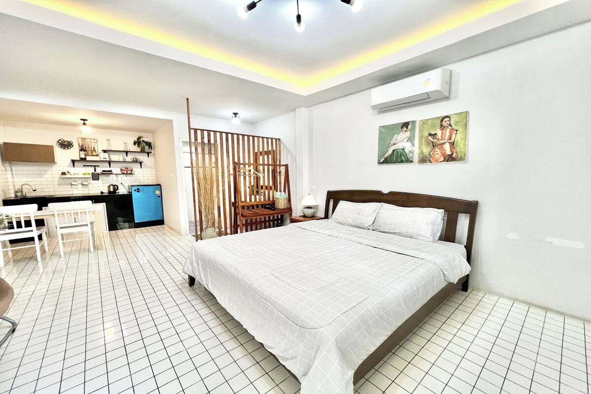 Quarto moderno com uma cama grande, uma escrivaninha e uma cadeira. Há uma área de cozinha com fogão e prateleiras.