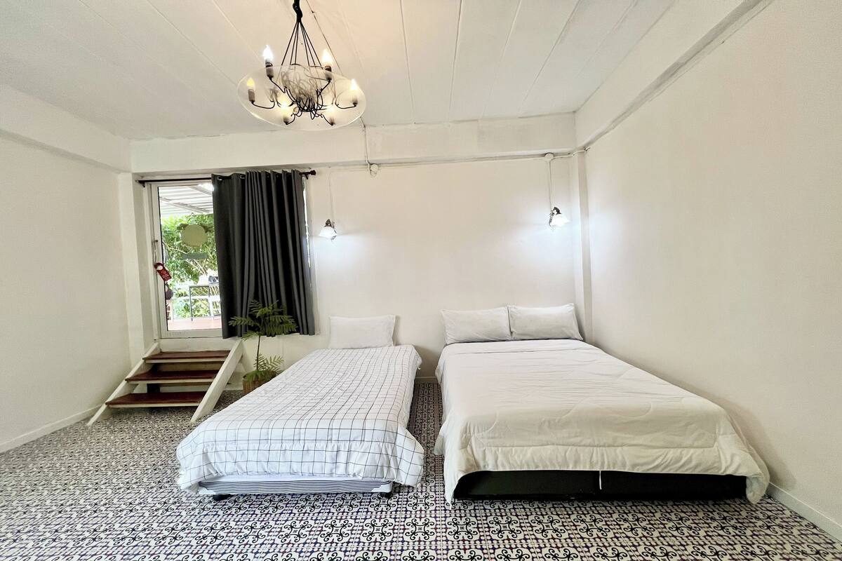 Quarto com duas camas, um candelabro, uma janela com cortinas e uma escadaria que dá para uma área externa.