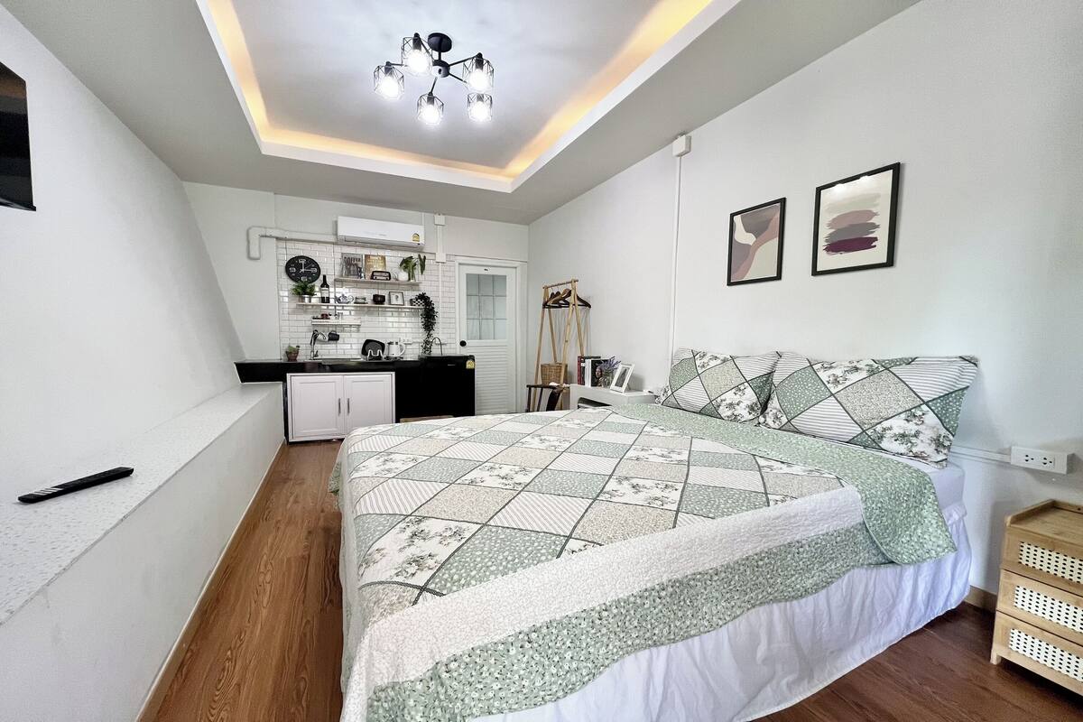 Quarto com cama, piso de madeira, parede branca, luminária no teto e mesa de cabeceira de madeira.