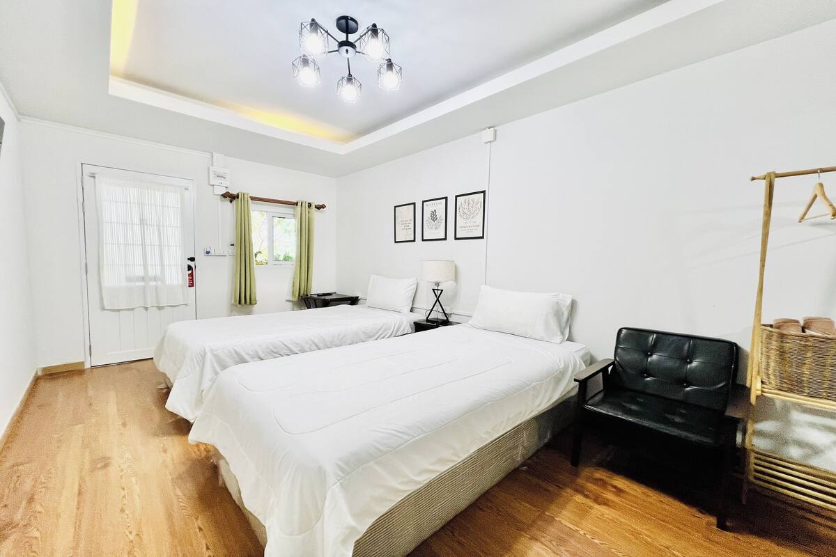 Quarto de hotel com duas camas, um sofá de couro preto, uma mesinha, uma luminária e uma prateleira fixada na parede.