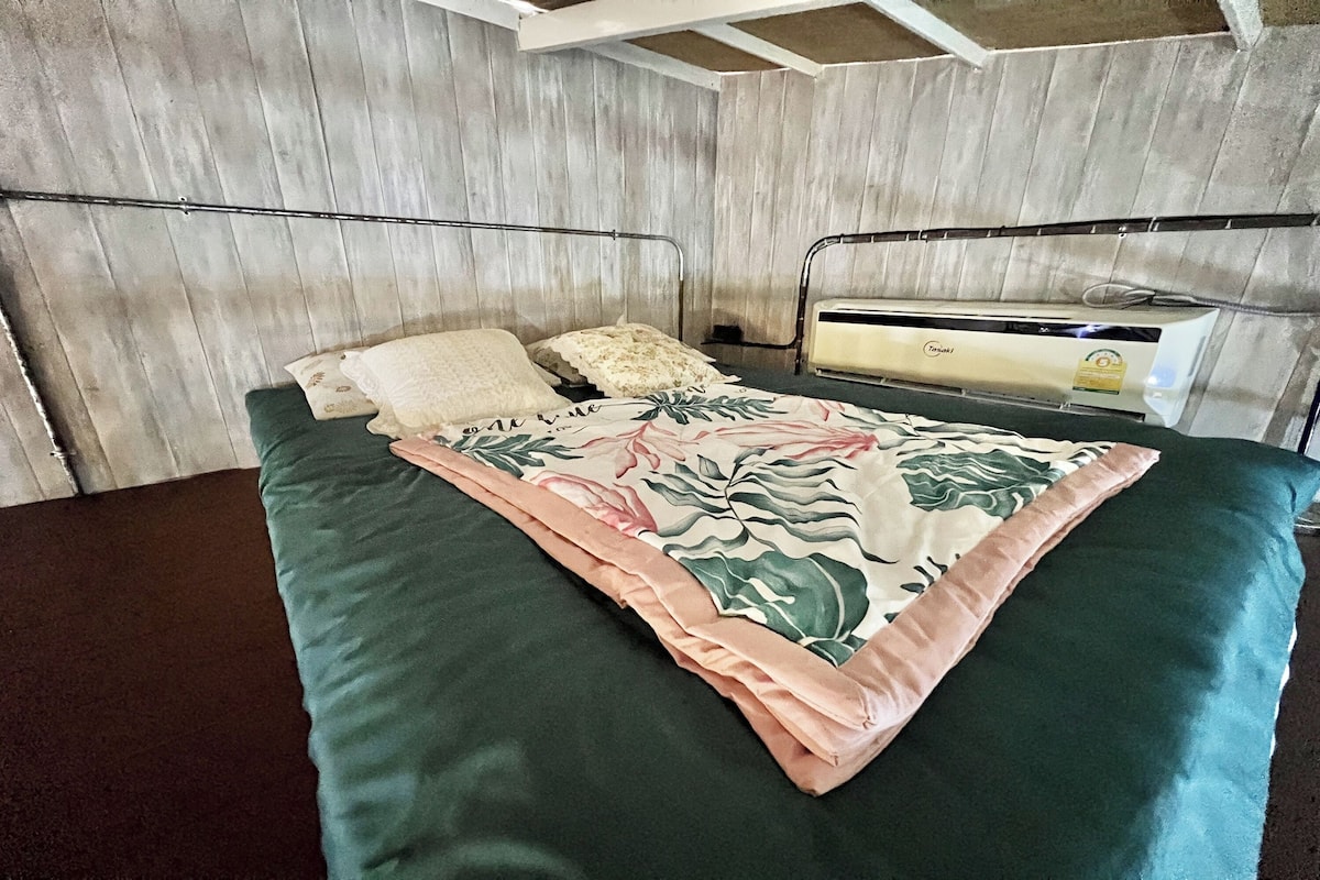 Cama com colcha florida e duas almofadas, ar condicionado fixado na parede e painel de parede de madeira.
