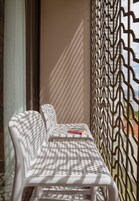 Uma varanda com duas cadeiras de vime brancas e uma mesa pequena, uma divisória metálica decorativa e vista para edifícios ao fundo.
