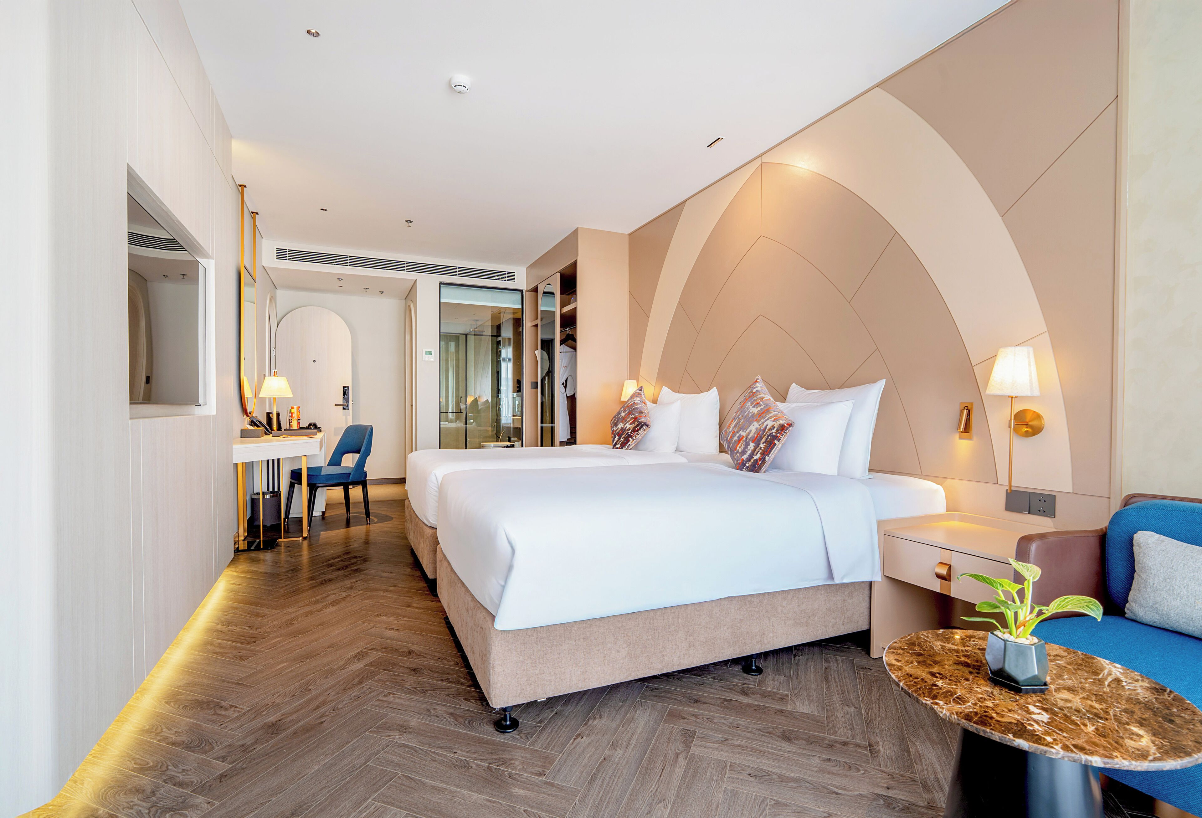 Quarto de hotel moderno com uma cama grande, uma escrivaninha com cadeira, um sofá e uma pequena mesa com uma planta.