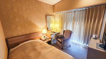 Quarto de hotel com uma cama grande, uma escrivaninha com luminária, uma cadeira e uma televisão sobre um suporte.
