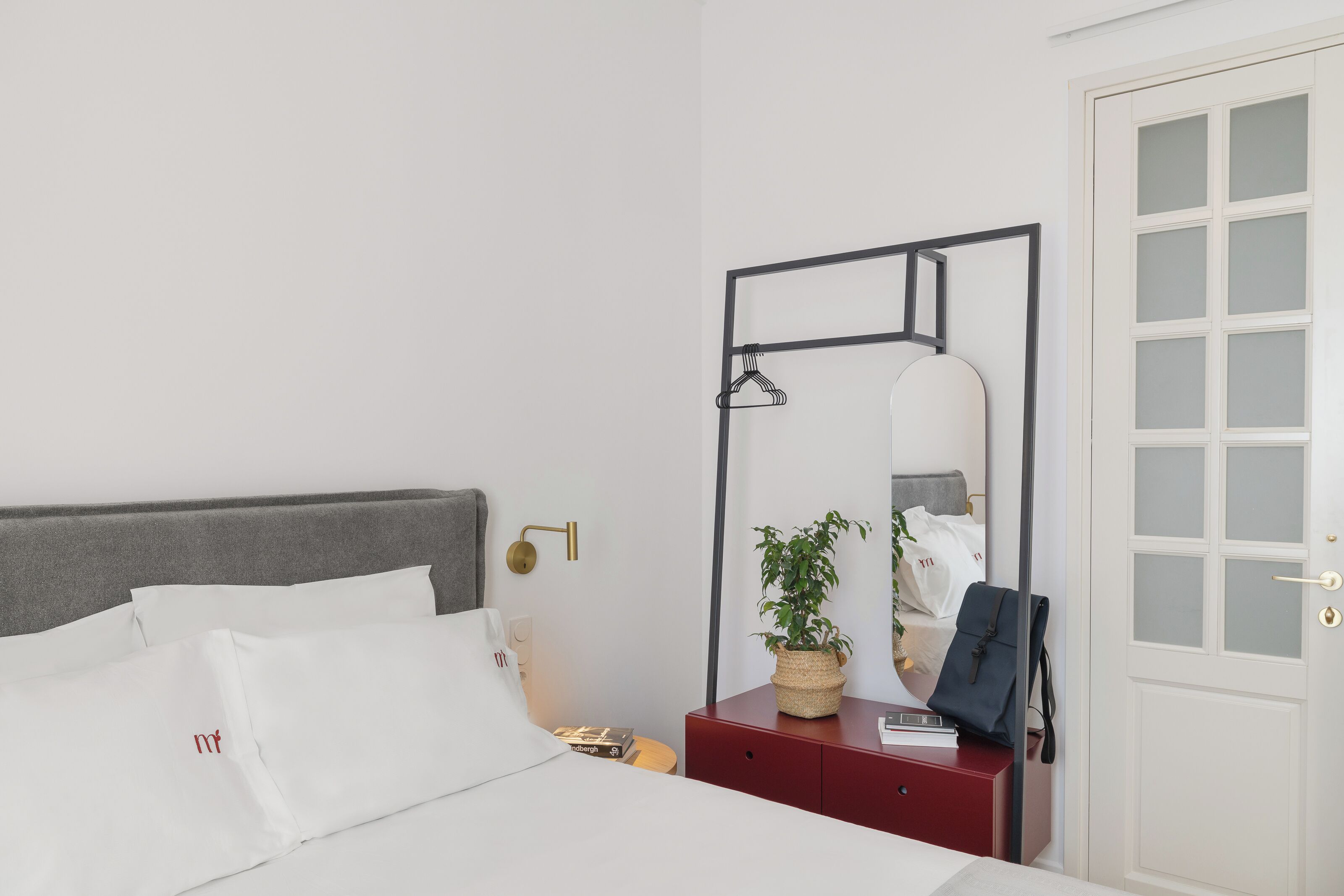 Cama bem arrumada com lençóis brancos, mesa de cabeceira com abajur, espelho, planta em vaso e porta com painéis de vidro fosco.