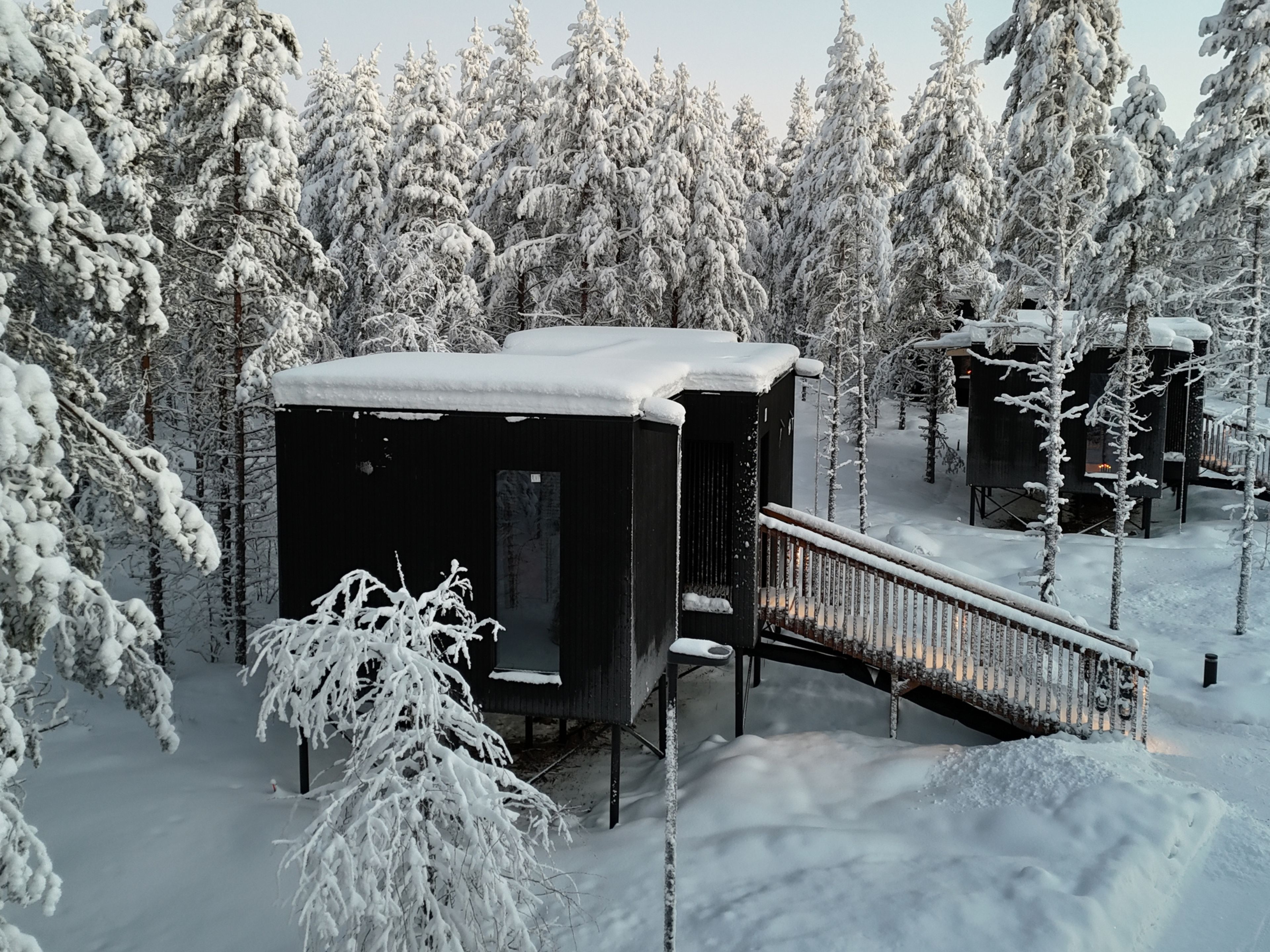 Uma cabana de madeira coberta pela neve, com varanda de madeira e escadas que levam à entrada.