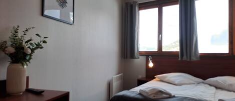 Quarto com cama, mesa de cabeceira, luminária, janela com cortinas e um quadro na parede.