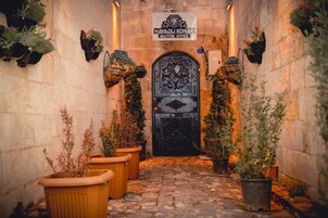 Um corredor de pedra estreito com uma porta decorativa, plantas em vasos e uma placa com a inscrição 'HAYADLI KONAK BÜTİK OTEL'.