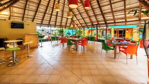 Uma ampla sala de jantar interna, com pé direito alto e iluminação natural, com várias mesas e cadeiras coloridas.
