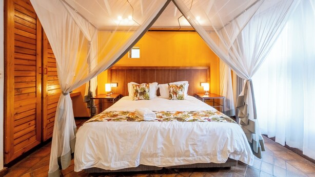 Quarto espaçoso com uma cama grande, cabeceira de madeira e um dossel. Há mesas de cabeceira com abajures e uma janela pequena.