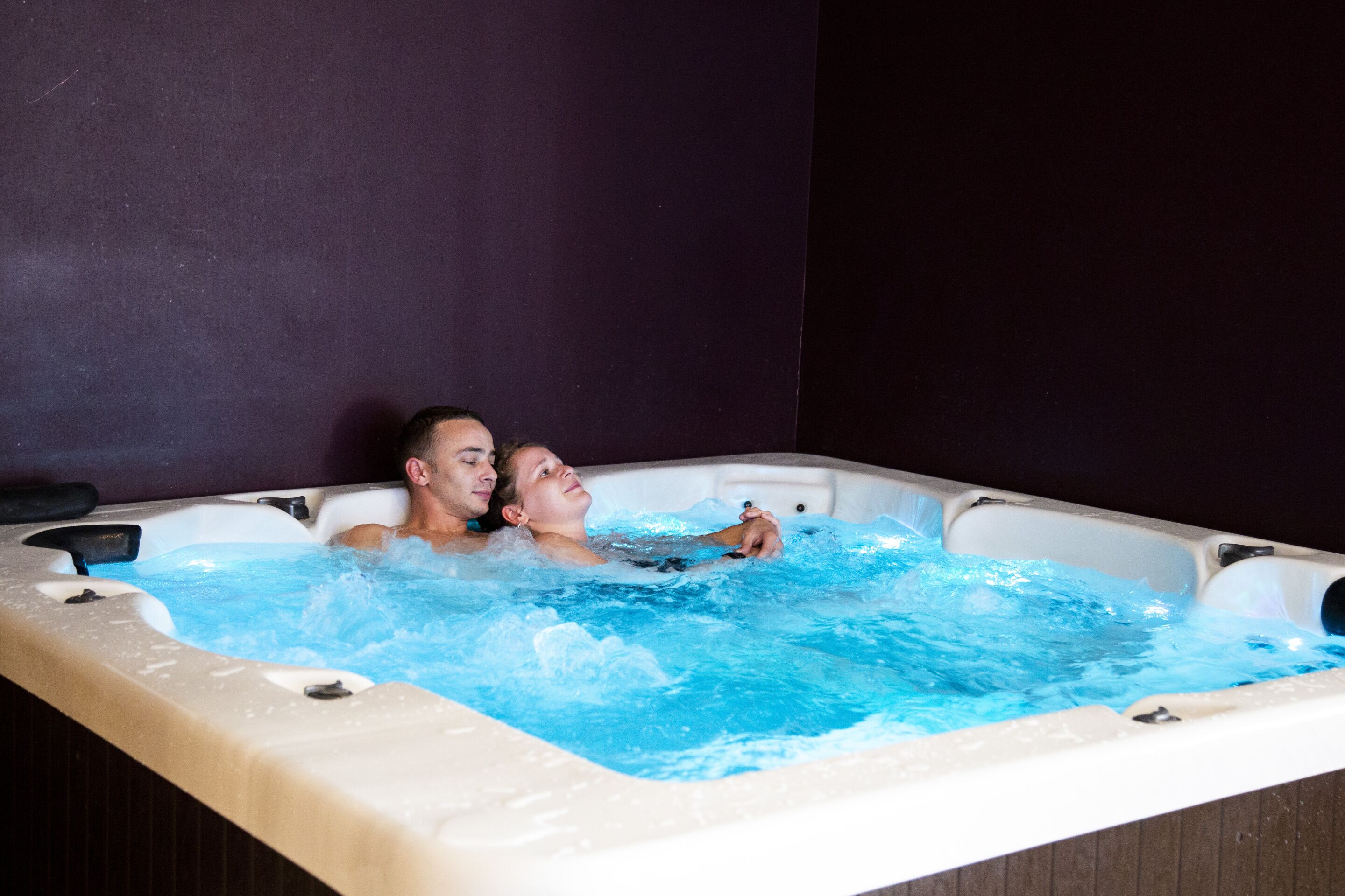 Um casal em uma banheira de hidromassagem com jatos, diante de uma parede escura.