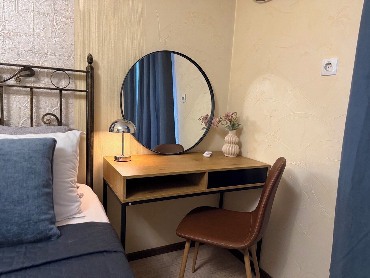 Quarto com cama, escrivaninha com cadeira, espelho redondo, abajur e um vaso com flores.