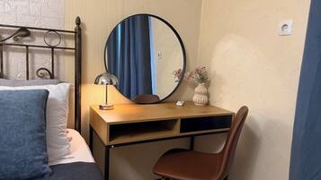 Quarto com cama, escrivaninha com cadeira, espelho redondo, abajur e um vaso com flores.