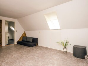 Quarto estilo loft com teto inclinado, sofá preto, planta em vaso e um banquinho cinza.