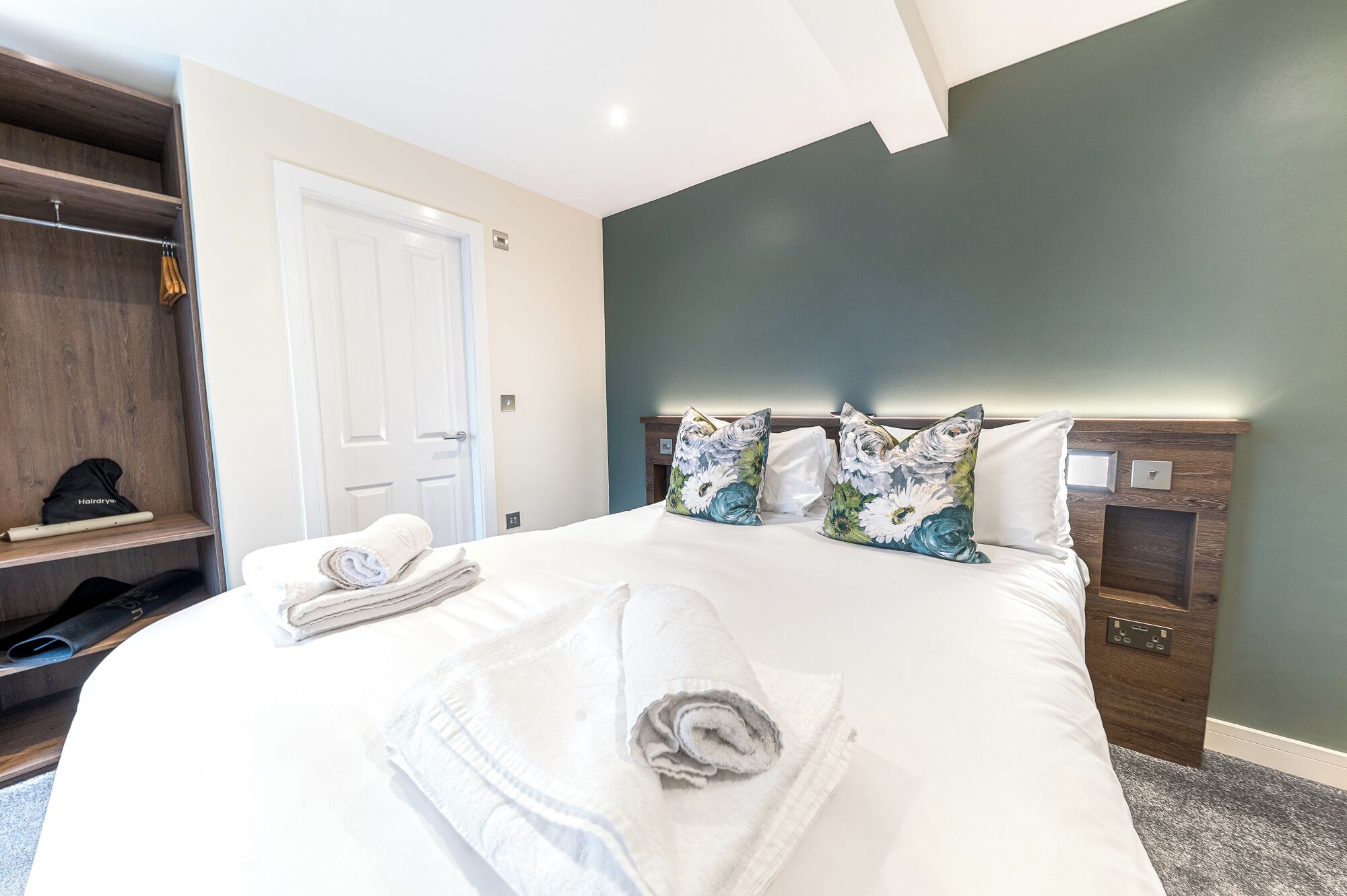 Quarto de hotel com cama, duas almofadas, cabeceira de madeira, porta branca e sapateira.