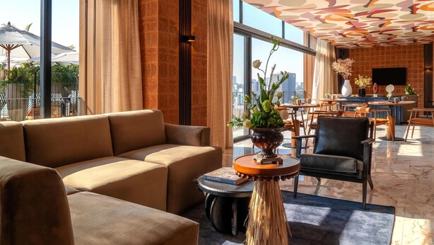 Um hall de hotel moderno com um sofá grande, uma mesa redonda com um vaso, e vista para a cidade através de amplas janelas.
