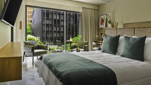 Quarto de hotel com uma cama grande, uma escrivaninha, uma cadeira, televisão e varanda com vista para o verde.