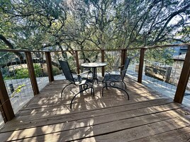 Um pátio externo com uma mesa e duas cadeiras, rodeado por um deck de madeira e guarda-corpo de metal.