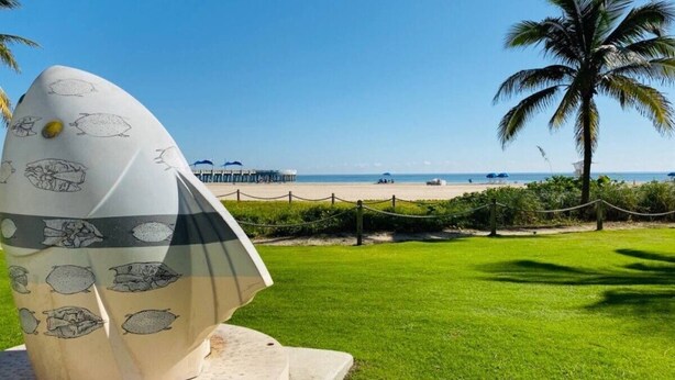 Uma grande escultura em forma de ovo com ilustrações de animais pré-históricos em um gramado, com vista para uma praia com palmeiras e um píer ao fundo.