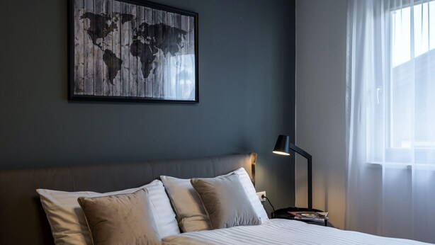 Uma cama bem arrumada com almofadas, um quadro de um mapa-múndi na parede, um abajur e uma janela com cortinas leves.